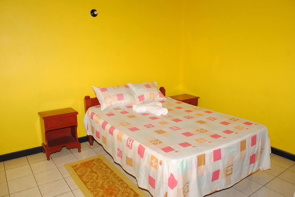 Uma cama com colcha estampada, duas mesas de cabeceira e um tapete floral num quarto com paredes amarelas.