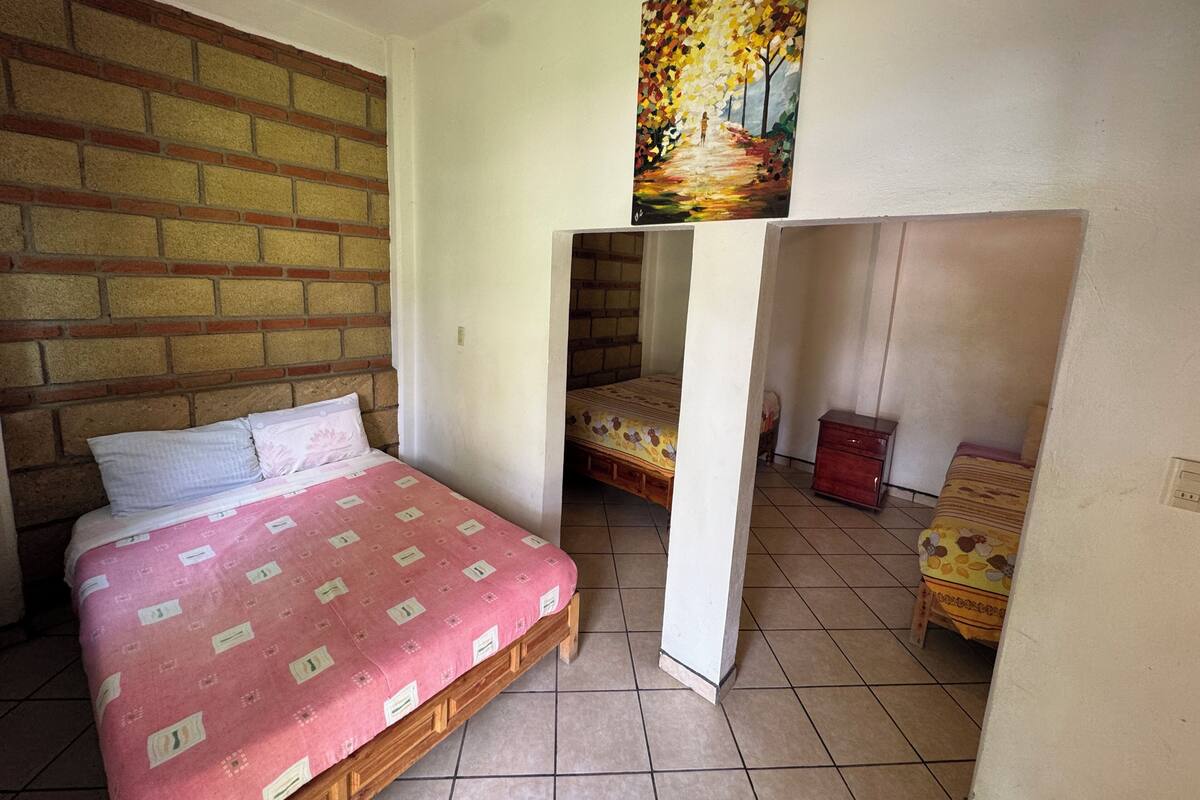 Quarto com parede de tijolos, cama com colcha estampada, quadro acima da cama e um pequeno armário de madeira.