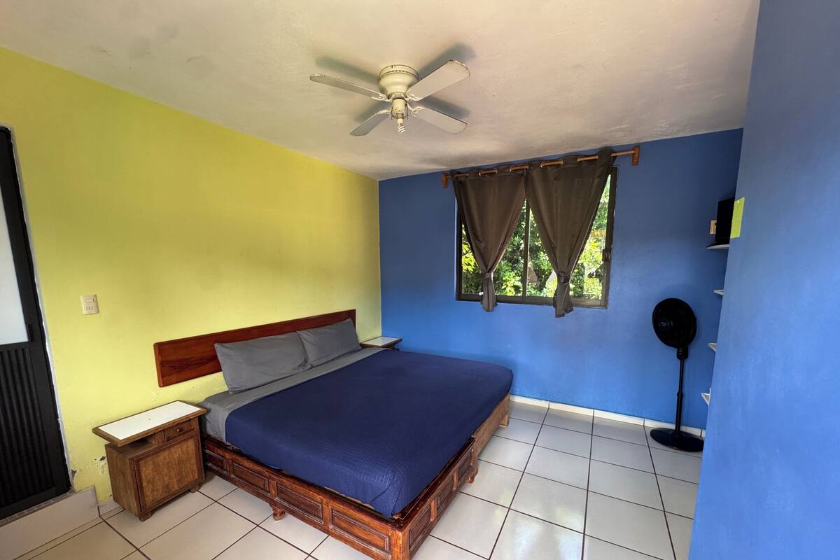 Quarto com cama, mesa de cabeceira, ventilador de teto, janela com cortinas e abajur preto.