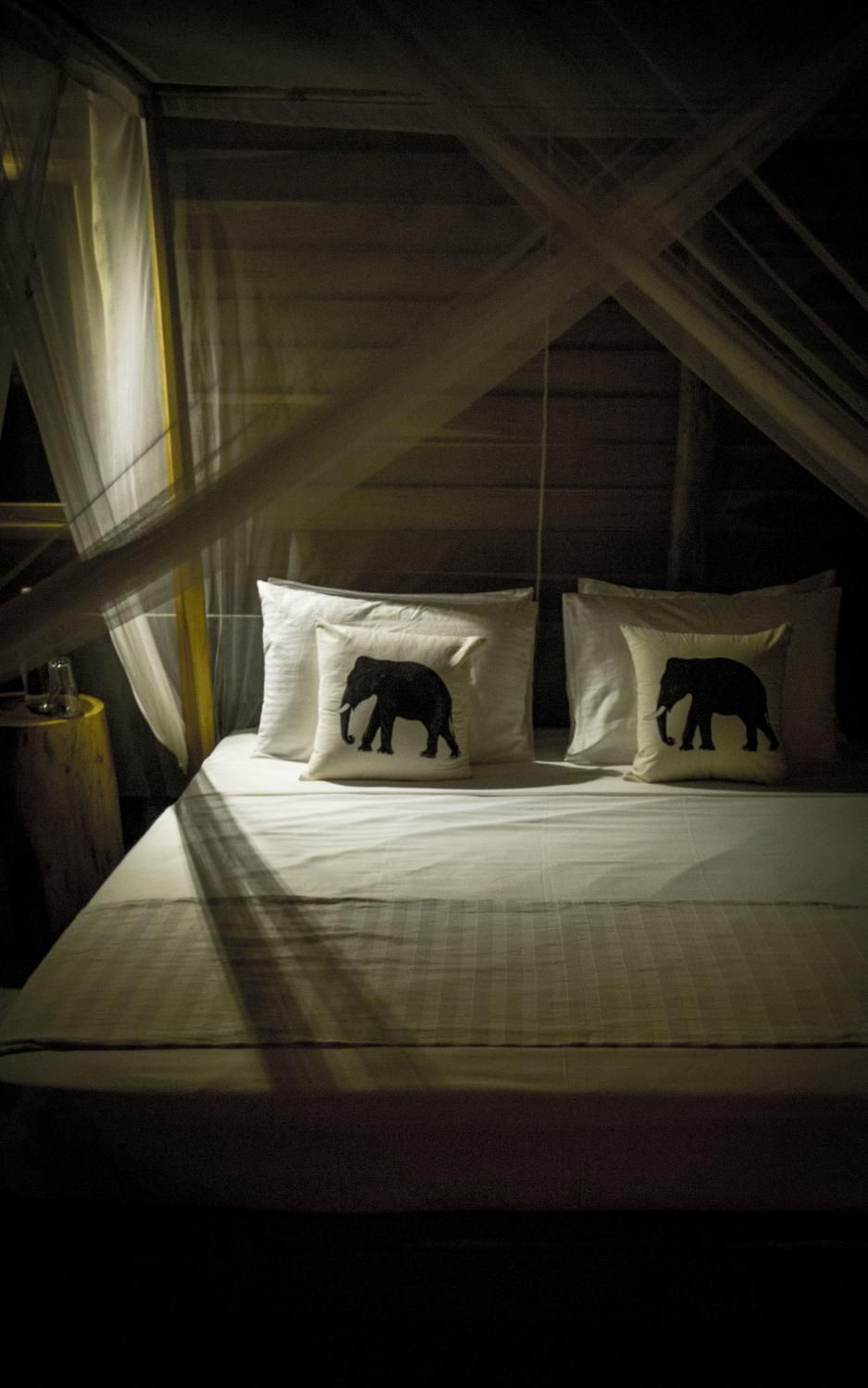 Cama com duas almofadas com silhuetas de elefantes, cabeceira de madeira e dossel.