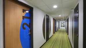 Corredor de hotel com carpete verde, portas numeradas e teto com iluminação embutida.