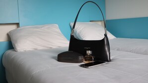 Cama bem arrumada com lençóis brancos, uma bolsa de mão preta, um frasco de perfume e um smartphone sobre a cama.