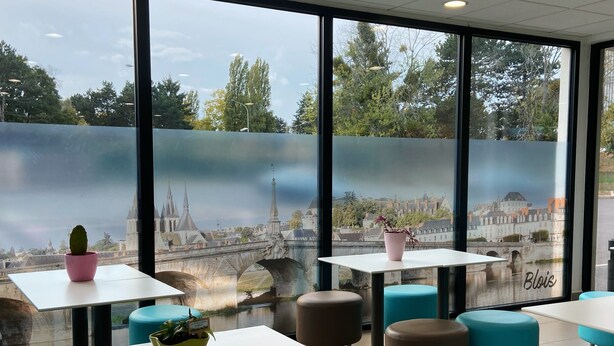 Uma sala de jantar moderna com mesas e cadeiras, janelas amplas com vista para a paisagem urbana e plantas decorativas em vasos.
