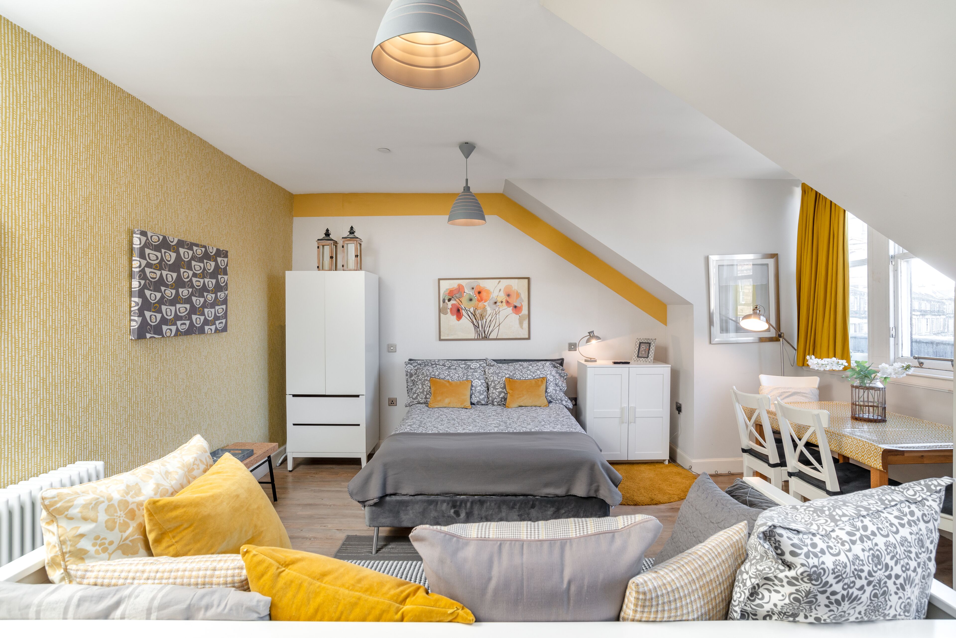 Quarto moderno com teto estilo loft, cama com roupa de cama estampada, cômoda branca e área de jantar com toalha de mesa amarela.