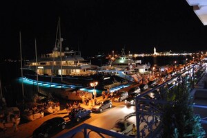 Uma marina à noite com barcos atracados, um edifício grande com varanda, e uma área movimentada à beira-mar com carros e pessoas.