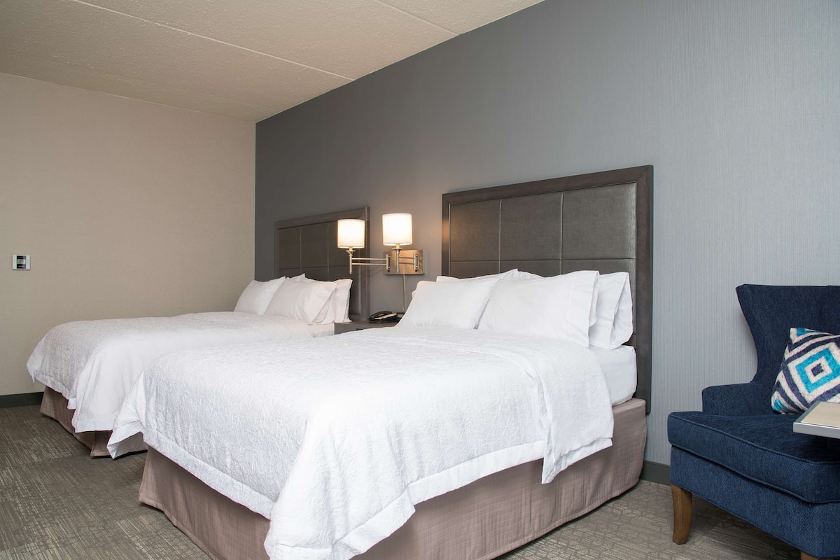 Quarto de hotel com duas camas, uma mesinha de cabeceira, uma luminária e uma cadeira.