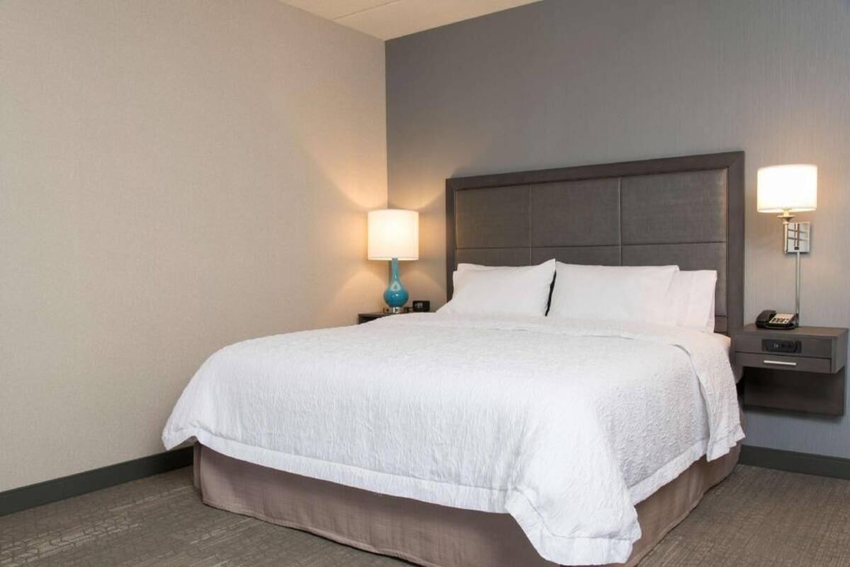 Quarto de hotel com uma cama grande, uma mesa de cabeceira com abajur e um telefone.