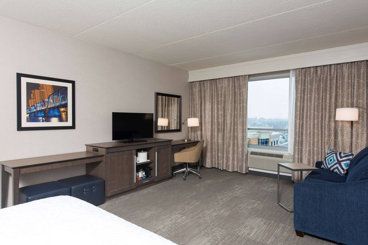 Quarto de hotel com uma cama, uma escrivaninha, uma cadeira, uma televisão e vista para a paisagem urbana através de grandes janelas.