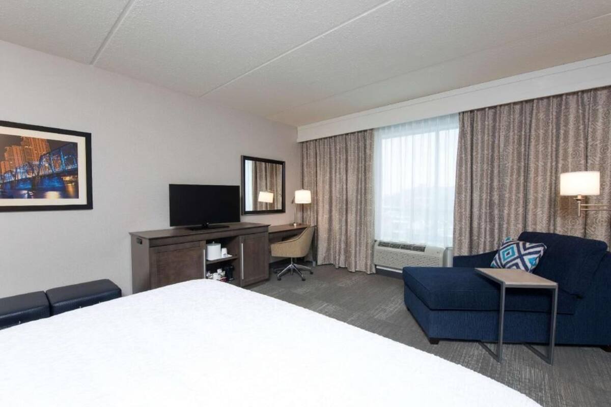 Quarto de hotel com cama, escrivaninha, poltrona, televisão, sofá e janela com cortinas.