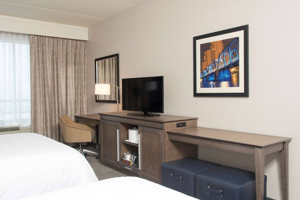 Quarto de hotel com uma cama, uma secretária com televisão, uma cadeira, abajur, quadro na parede e janela com cortinas.