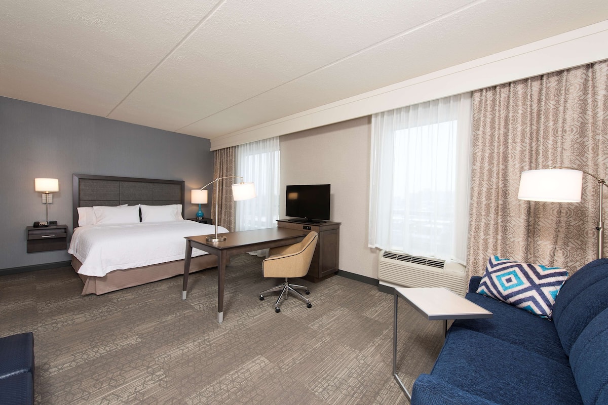 Quarto de hotel com cama, escrivaninha, cadeira, televisão e sofá.