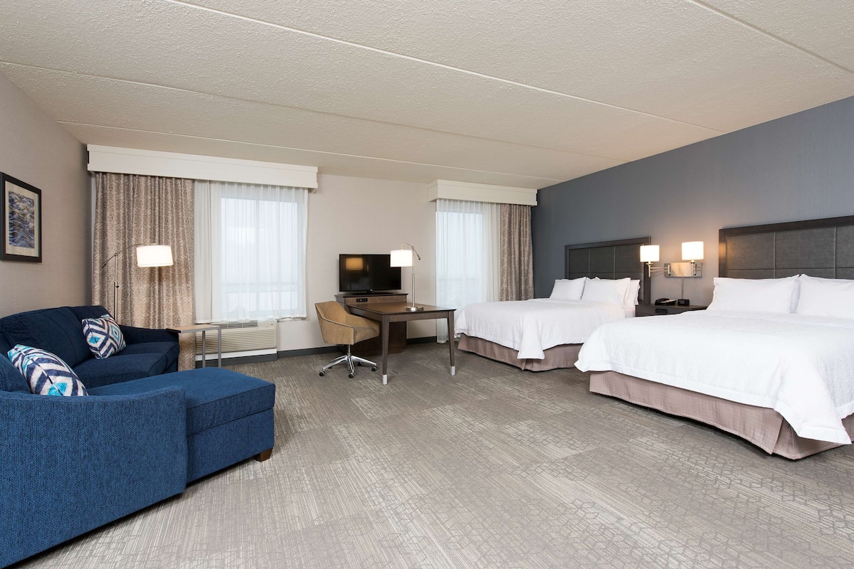 Quarto de hotel com sofá, duas camas, escrivaninha, televisão e luminária.