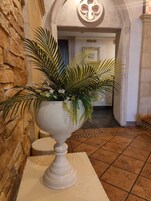 Urna branca com arranjo floral sobre mesa de pedestal em corredor com paredes de pedra e chão de azulejos.