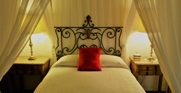 Uma cama com colcha branca, um travesseiro vermelho e uma cabeceira metálica decorativa. Há duas mesas de cabeceira com luminárias e uma mesinha com um objeto decorativo.