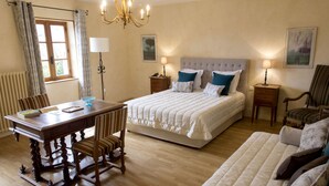 Quarto com cama de casal, escrivaninha de madeira, cadeira, mesa de cabeceira, abajur, radiador e janela com cortinas.