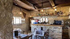 Um espaço de bar rústico, com paredes de pedra, vigas de madeira e diversas garrafas e copos nas prateleiras.