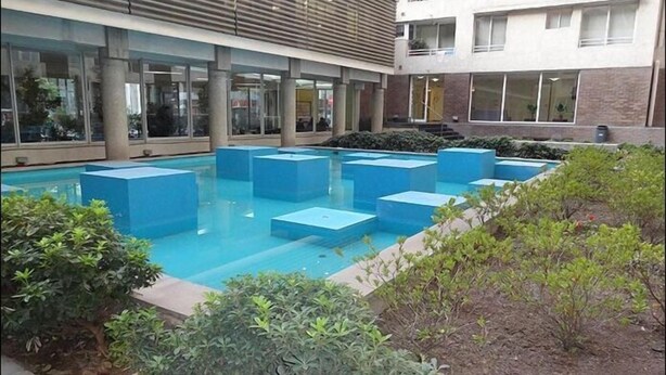Piscina com degraus azuis retangulares, rodeada por vegetação e um edifício com janelas amplas e fachada de tijolos.