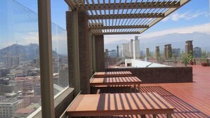 Terraço no telhado com mesas e cadeiras de madeira, uma pérgula e vista para a cidade.