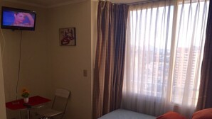Quarto de hotel com uma cama, uma secretária com cadeira, uma televisão fixada na parede e uma janela com cortinas.