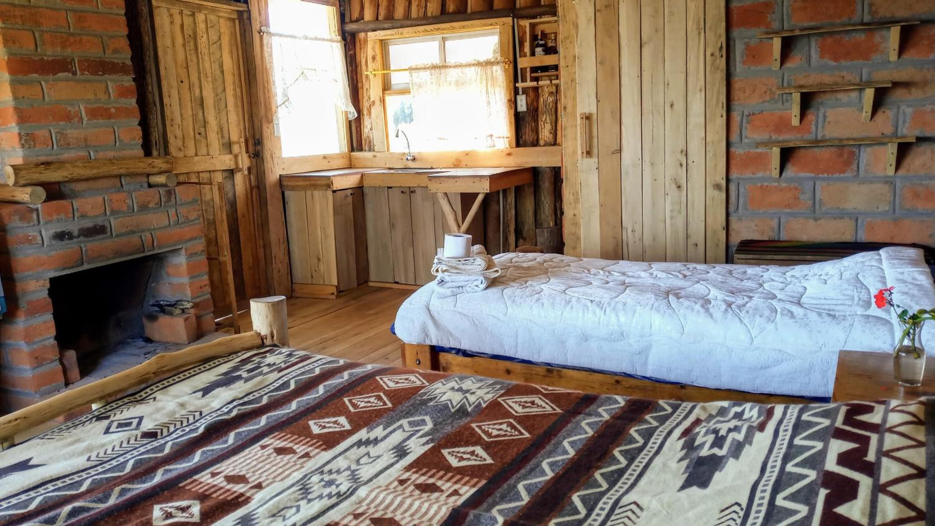 Quarto rústico com lareira de tijolos, uma mesa de madeira, cama com manta estampada e janela com cortinas.