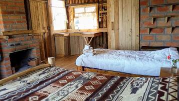 Quarto rústico com lareira de tijolos, uma mesa de madeira, cama com manta estampada e janela com cortinas.