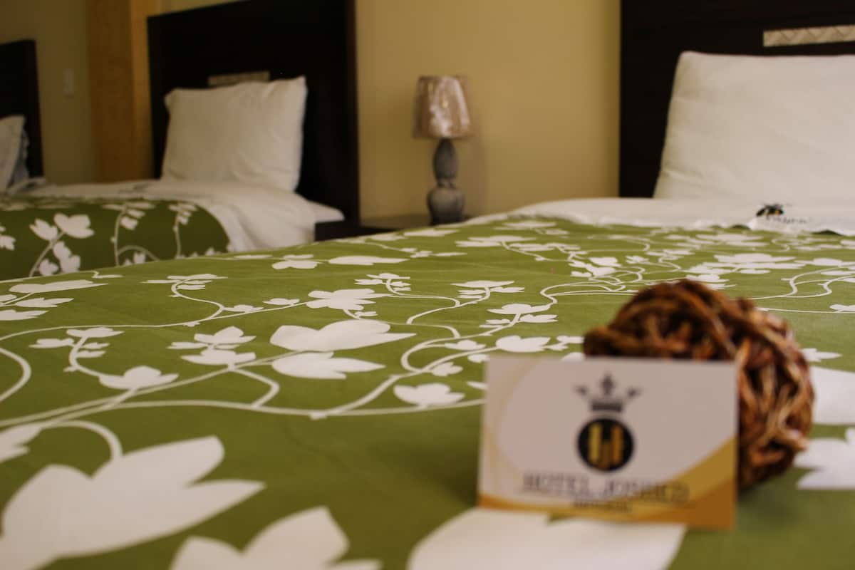 Quarto de hotel com cama coberta por roupa de cama estampada com flores verdes, cabeceira de madeira, mesa de cabeceira com abajur e placa com o nome do hotel.