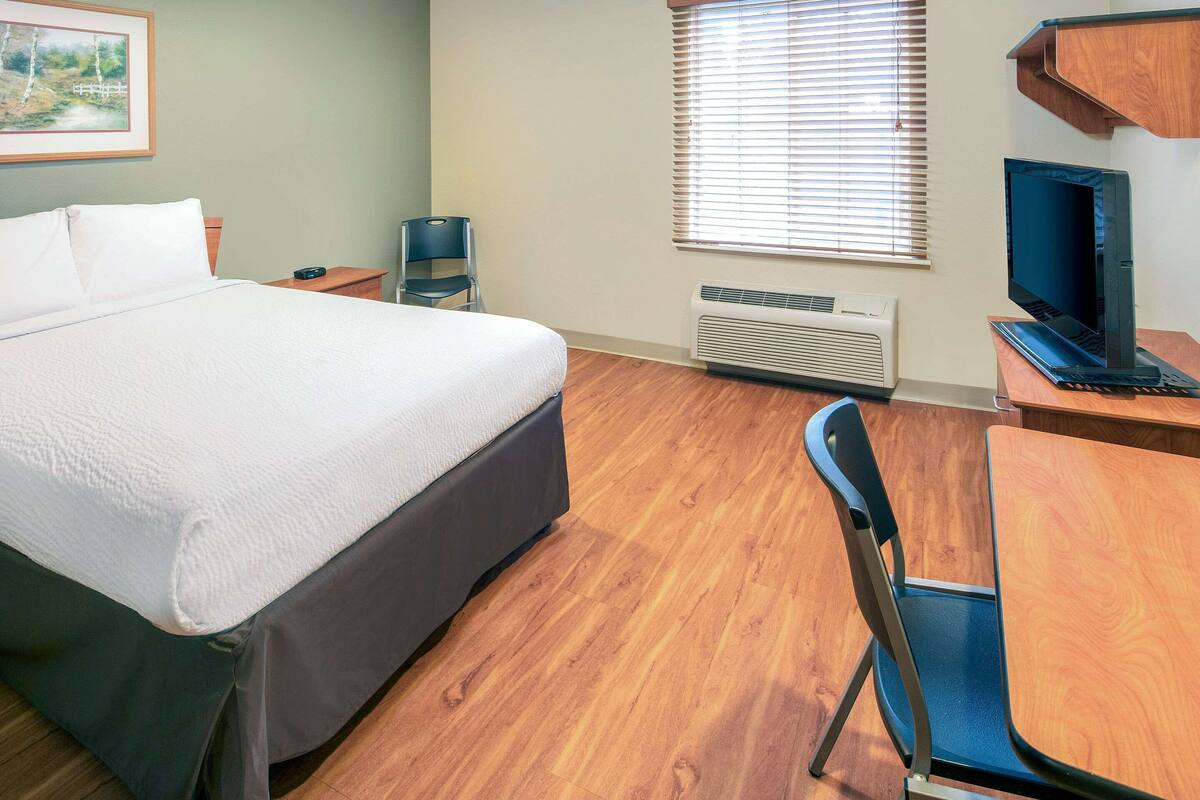 Quarto de hotel com cama, escrivaninha, cadeira, televisão e janela com persianas.