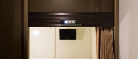 Quarto de hotel com uma cama de solteiro, uma televisão pequena e uma cortina.