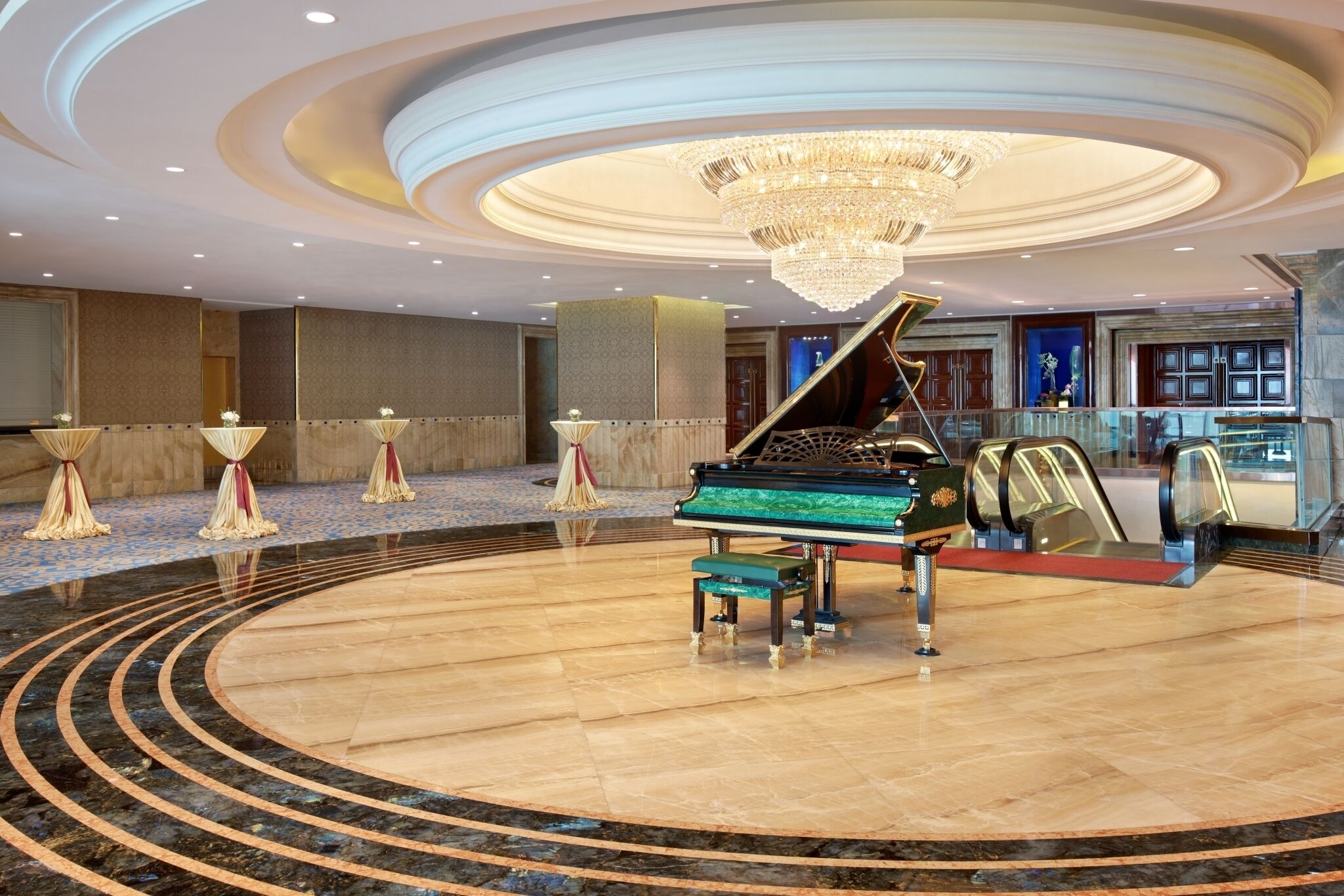Um piano de cauda imponente num palco circular, com um candelabro acima, rodeado por um chão de mármore polido e um guarda-corpo de vidro.