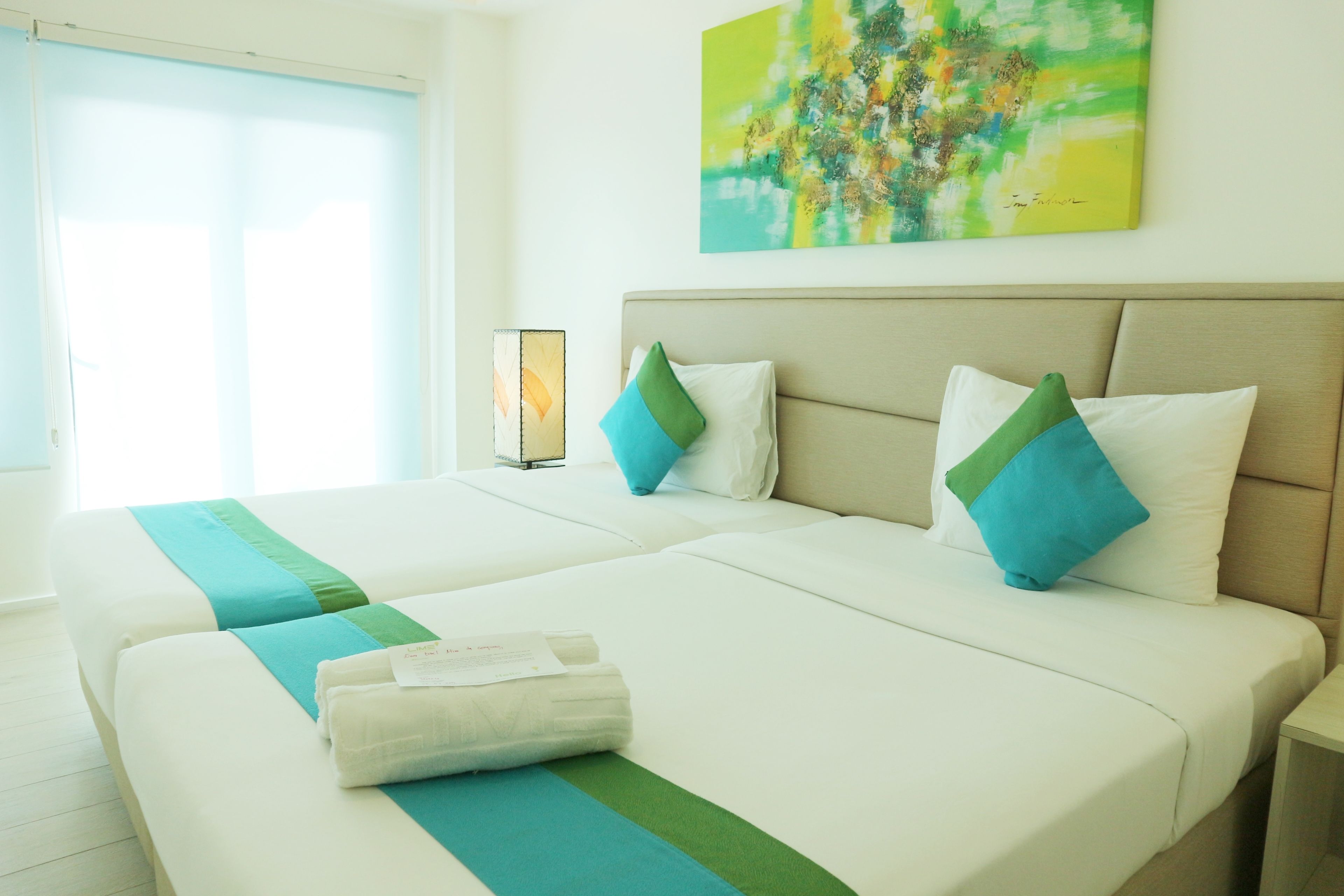 Quarto de hotel com duas camas, cada uma com colcha listrada branca e verde, cabeceira bege e travesseiro azul. Há uma pintura na parede acima das camas e uma mesa de cabeceira com abajur ao lado de cada cama.