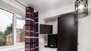 Um quarto pequeno com janela, cortina, televisão e armário.