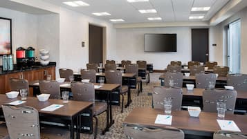 Uma sala de conferência com várias mesas e cadeiras, equipada com máquinas de café, copos de água e uma televisão de tela plana na parede.