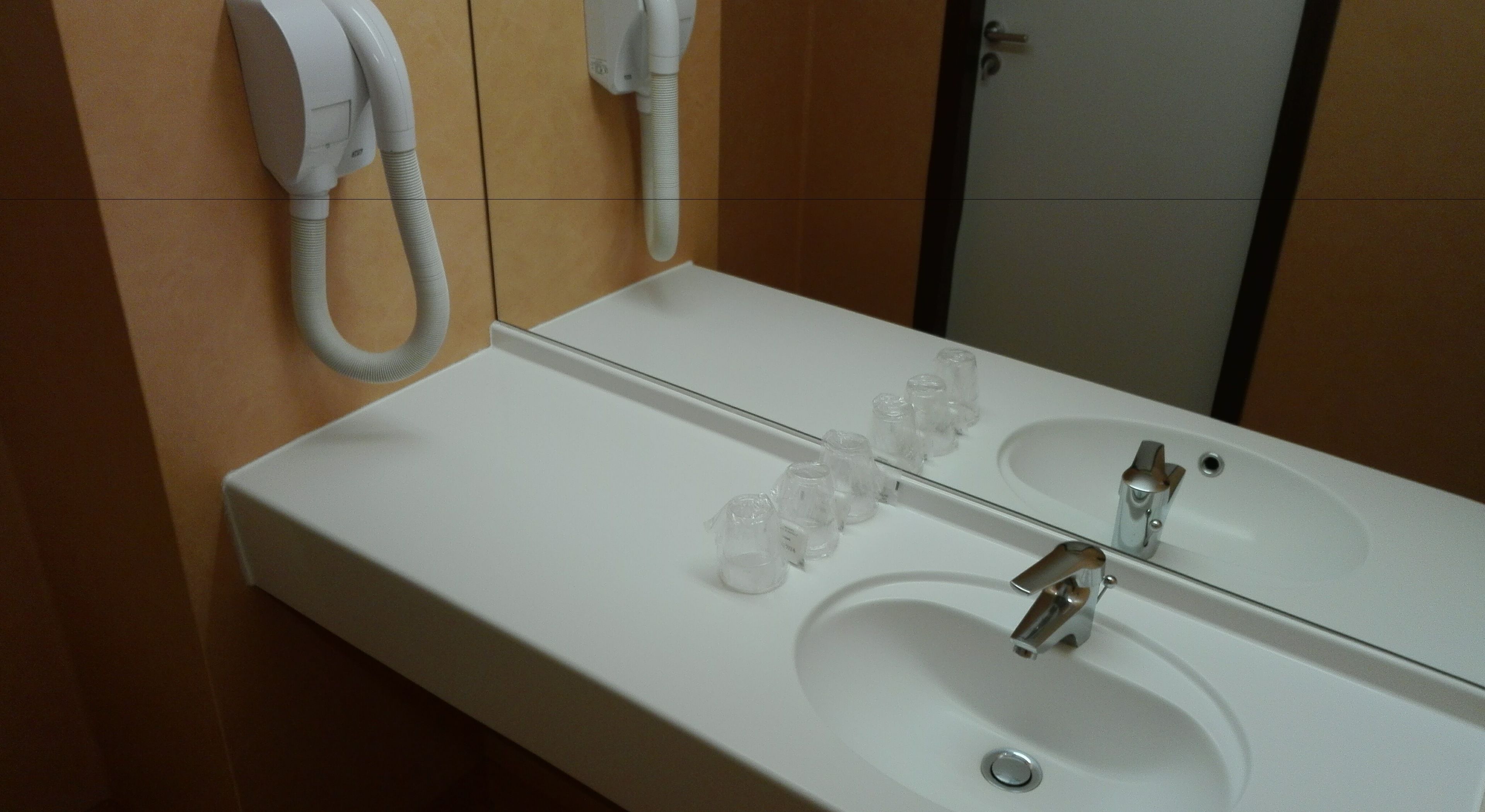 Casa de banho com lavatório branco, duas espelhas e secador de cabelo na parede.