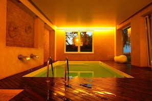Uma piscina interior moderna com deck de madeira, parede de tijolo e janela com vista para árvores.