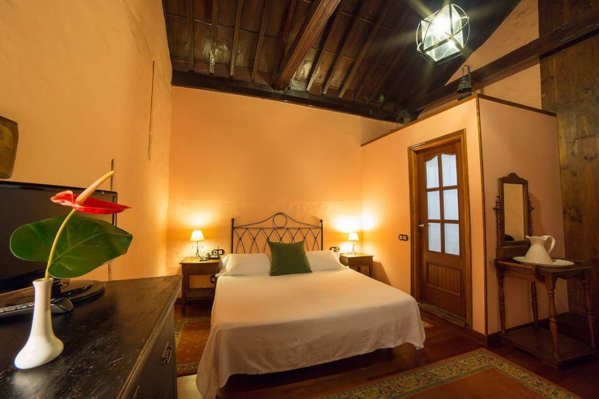 Quarto aconchegante com teto de madeira, cama com roupa de cama branca, duas luminárias de cabeceira e mesa de cabeceira com um vaso de flores.