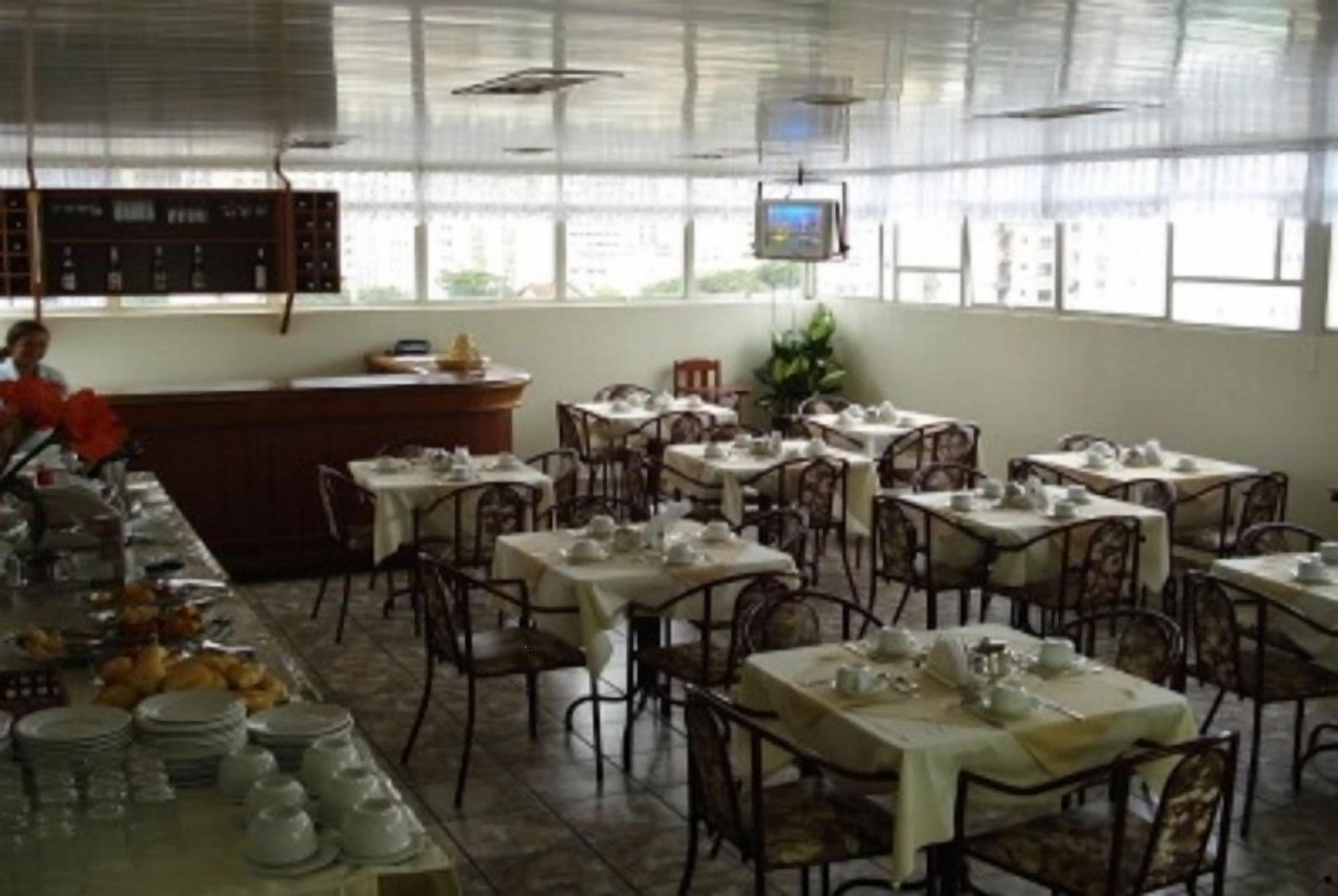Uma área de refeições com várias mesas preparadas para o serviço, um balcão com um quadro de menu e uma grande janela que proporciona luz natural.