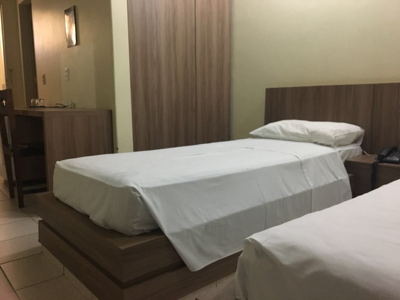 Quarto de hotel com duas camas, cabeceira de madeira, mesa de cabeceira com telefone e porta com espelho.