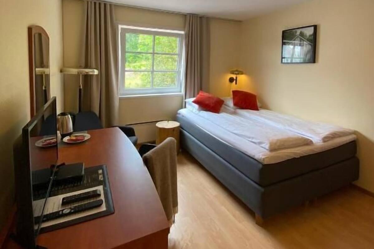 Quarto de hotel com escrivaninha, cadeira, cama, mesa de cabeceira, luminária e janela com cortinas.