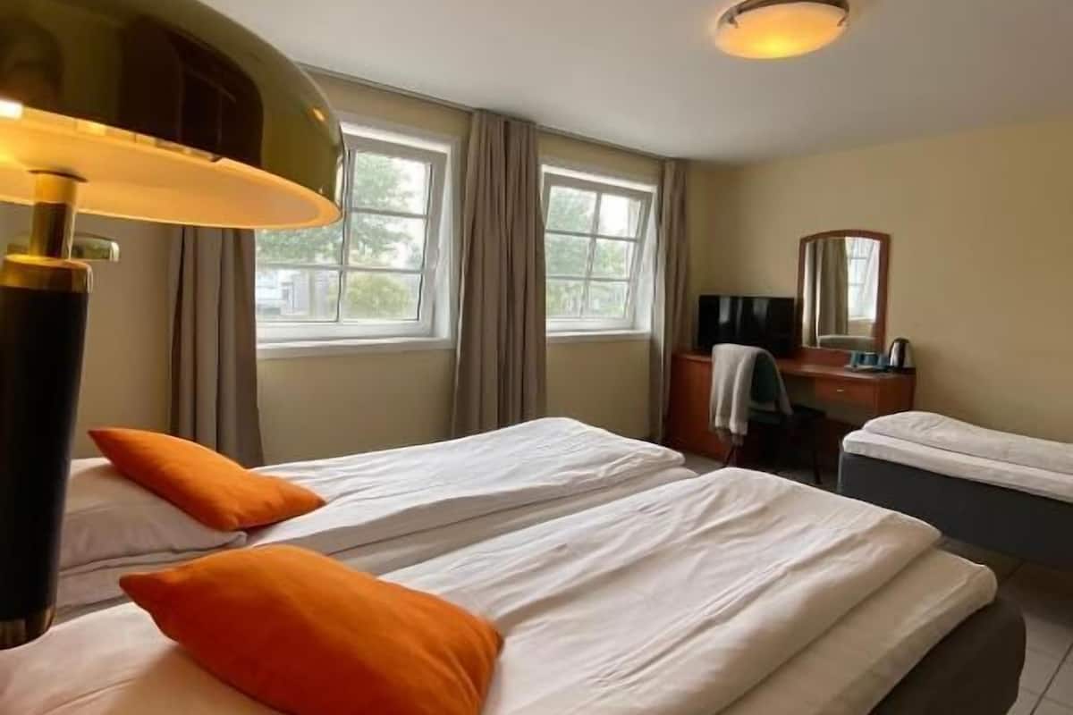 Quarto de hotel com duas camas, uma escrivaninha, uma cadeira, um abajur e uma luminária de teto.