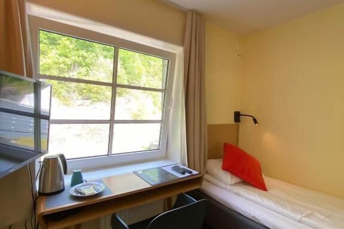 Um quarto pequeno com uma escrivaninha, uma cadeira e uma cama. Há uma janela com cortinas, um abajur e um travesseiro vermelho na cama.