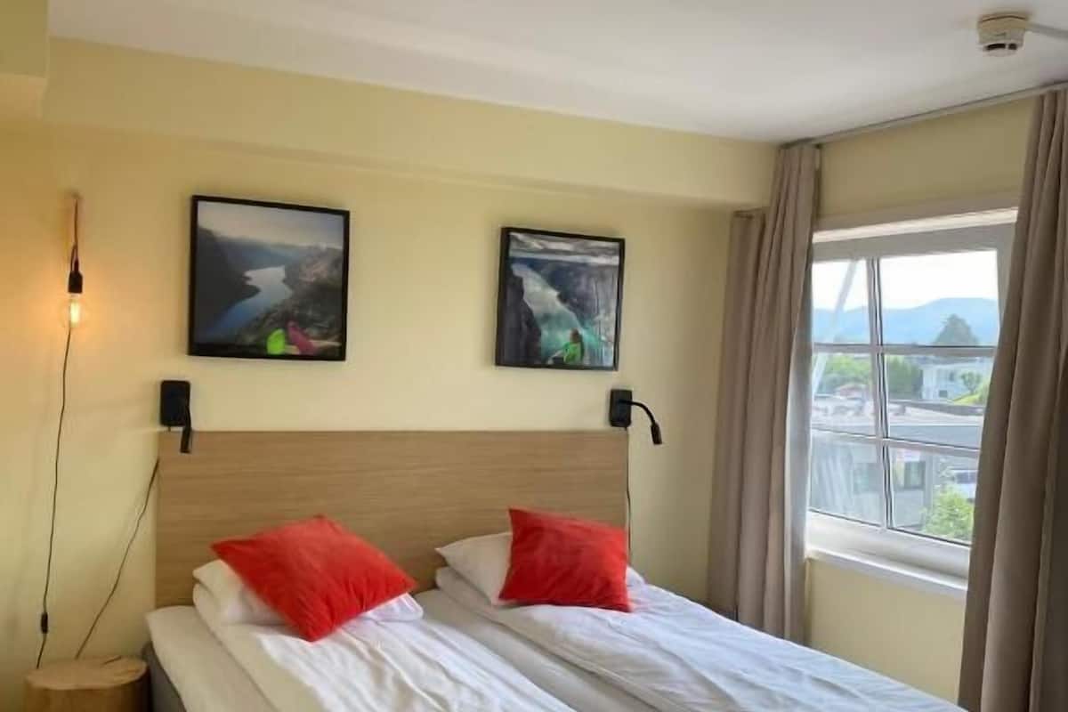 Uma cama de casal com roupa de cama vermelha e branca, dois quadros na parede, um abajur e uma janela com cortinas.