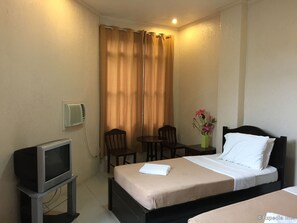 Quarto de hotel com cama, televisão, escrivaninha, duas cadeiras e janela com cortinas.