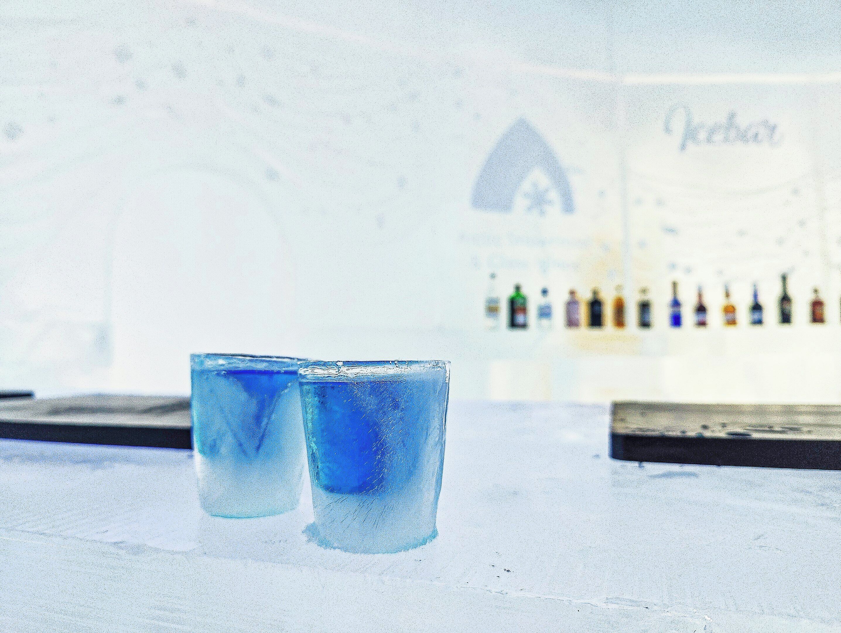 Dois copos de líquido azul sobre uma mesa, com um fundo desfocado de um bar e garrafas.