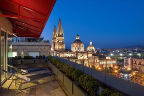 Terraço no telhado com espreguiçadeiras, um toldo vermelho e vista para edifícios históricos ao entardecer.