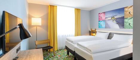 Quarto de hotel com duas camas, uma escrivaninha com luminária, uma cadeira, televisão e uma janela grande com cortinas.