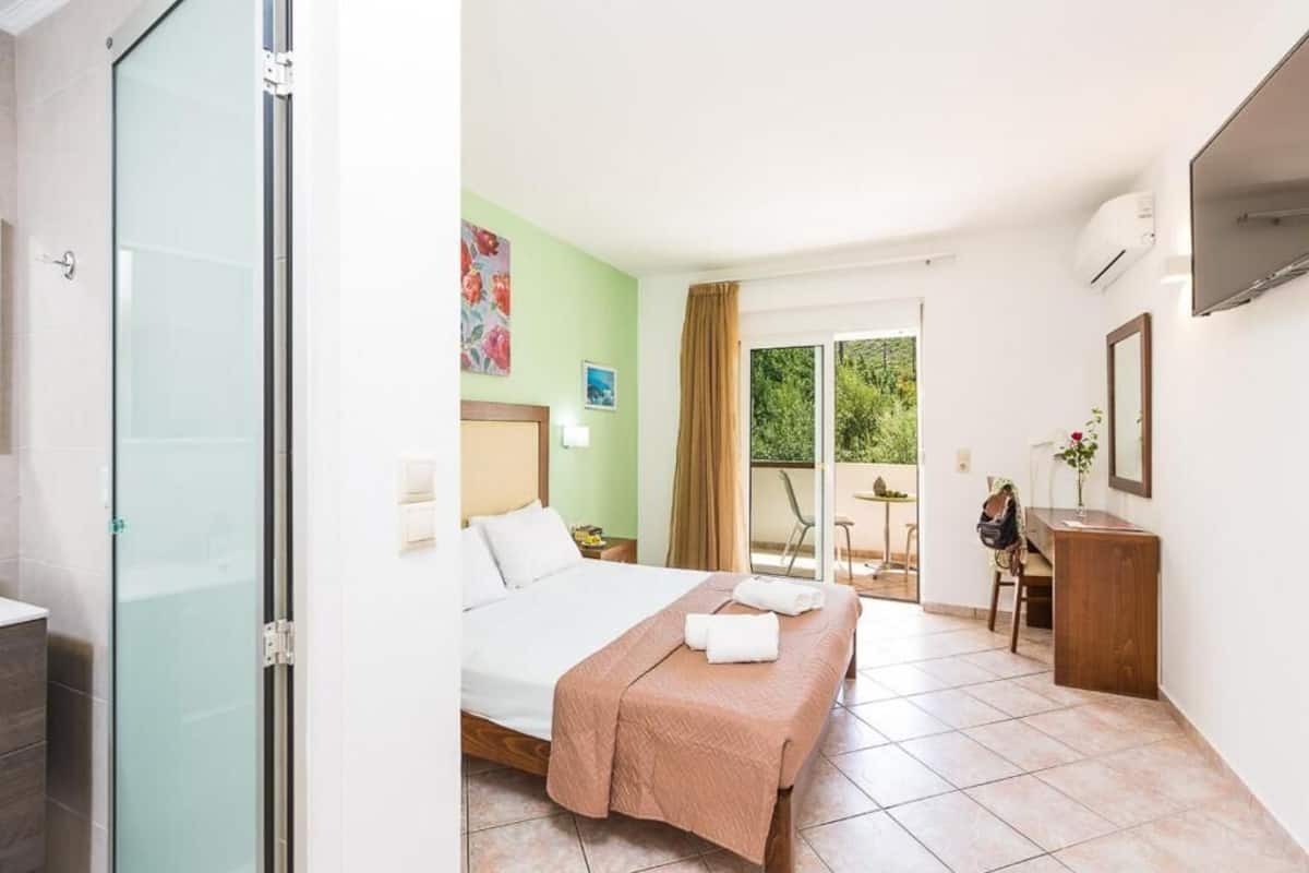 Quarto de hotel com cama, escrivaninha, cadeira, banheiro e varanda com vista para o verde.