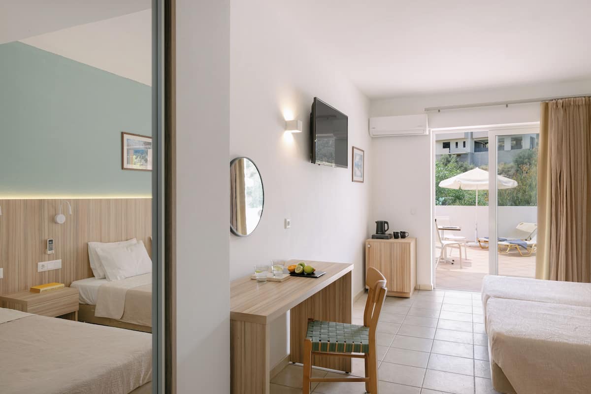 Quarto de hotel com duas camas, uma escrivaninha com cadeira, televisão e varanda com mobiliário.