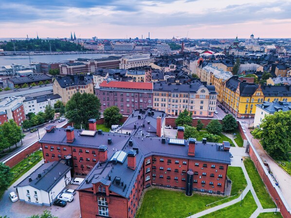 Um edifício grande e histórico, com várias alas, rodeado por um extenso jardim bem cuidado e um rio ao fundo.