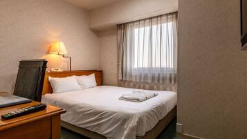 Quarto de hotel com uma cama, uma escrivaninha com abajur, um controle remoto e uma janela com cortinas leves.