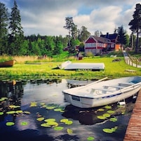 Um lago tranquilo com um barco atracado à direita, cercado por vegetação exuberante e uma casa vermelha ao fundo.
